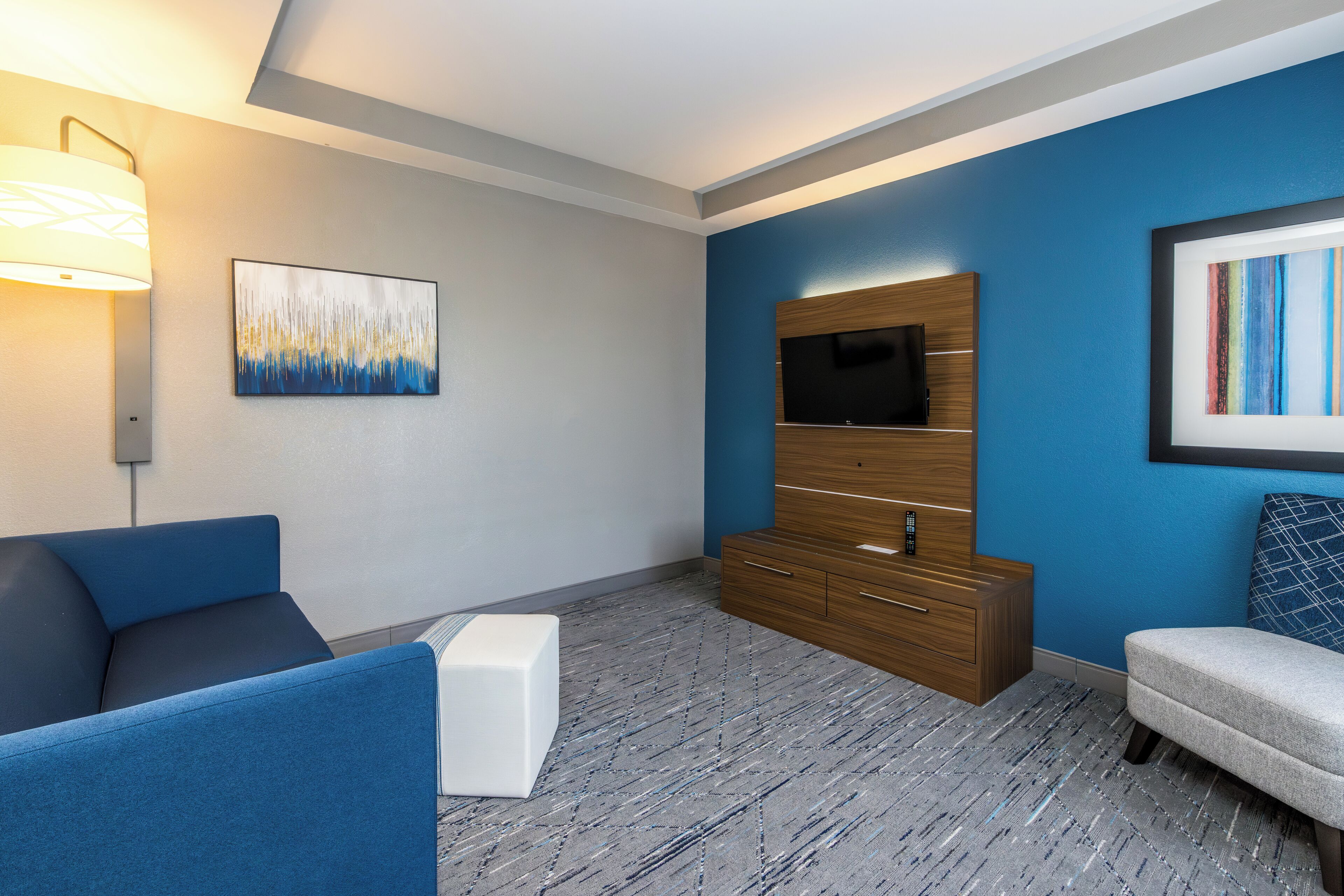 Quarto de hotel moderno com parede azul de destaque, televisão de tela plana presa a um armário de madeira, mesa de cabeceira branca e sofá azul.