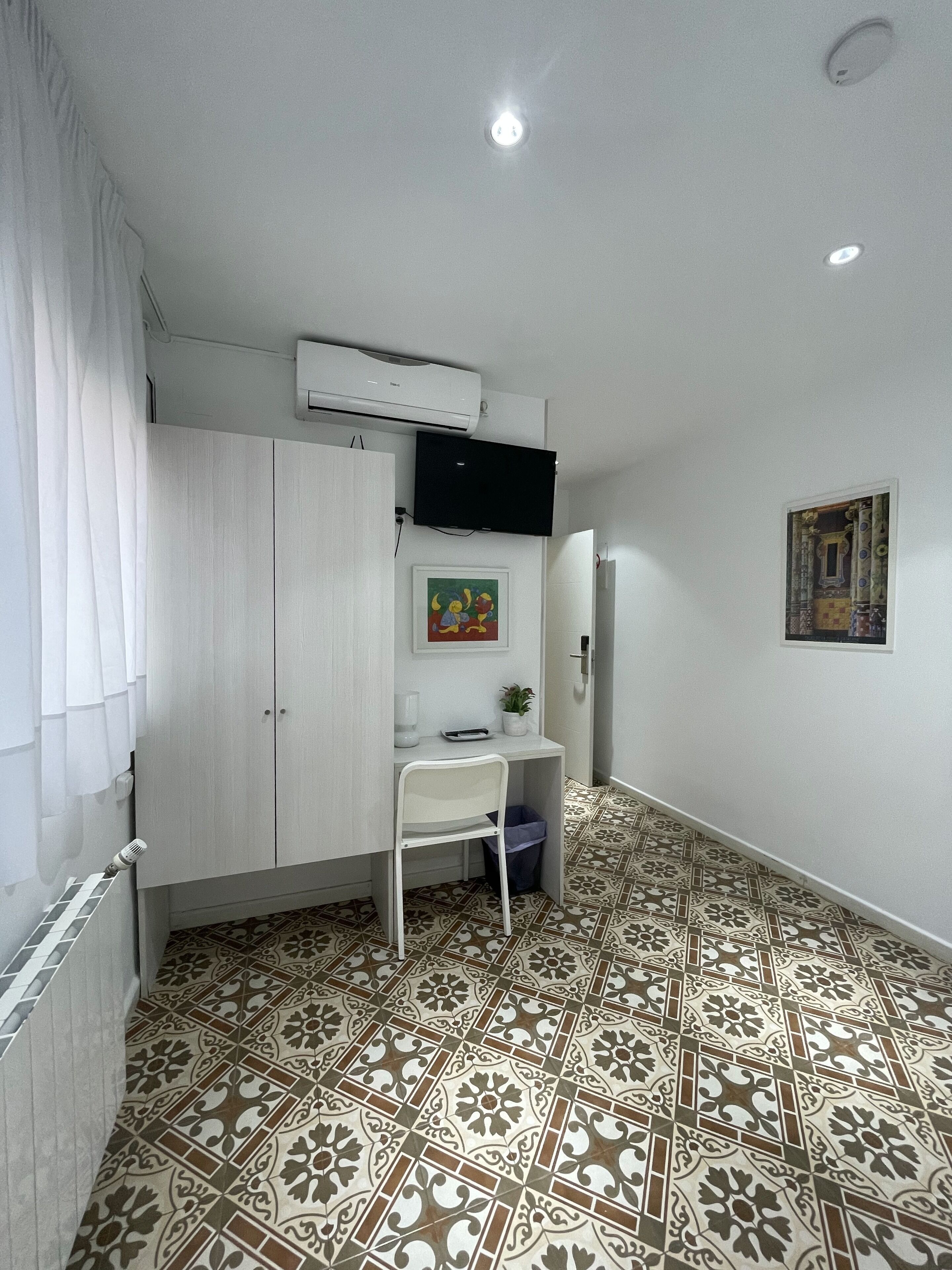 Quarto com guarda-roupa branco, escrivaninha com cadeira, televisão de tela plana montada acima da escrivaninha, uma pequena planta na escrivaninha, chão estampado e um quadro na parede.