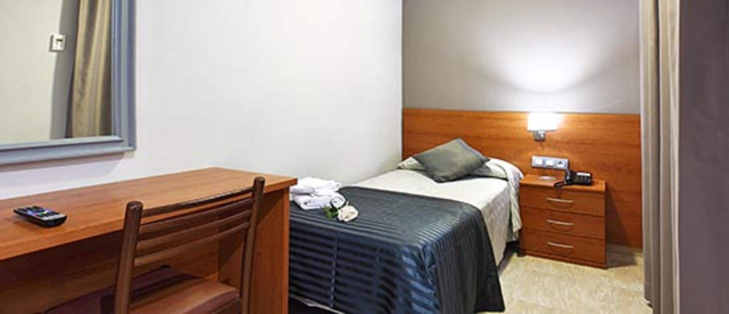 Quarto de hotel com escrivaninha de madeira, cama com colcha azul escura, mesa de cabeceira com abajur e cómoda de madeira.