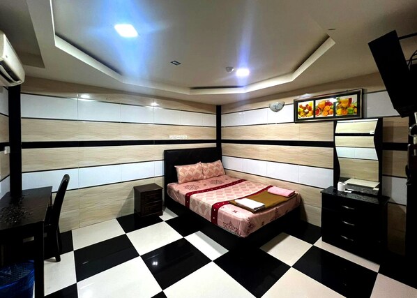 Um quarto com piso quadriculado em preto e branco, uma cama com colcha estampada em rosa e branco, uma escrivaninha com cadeira e uma TV fixada na parede.