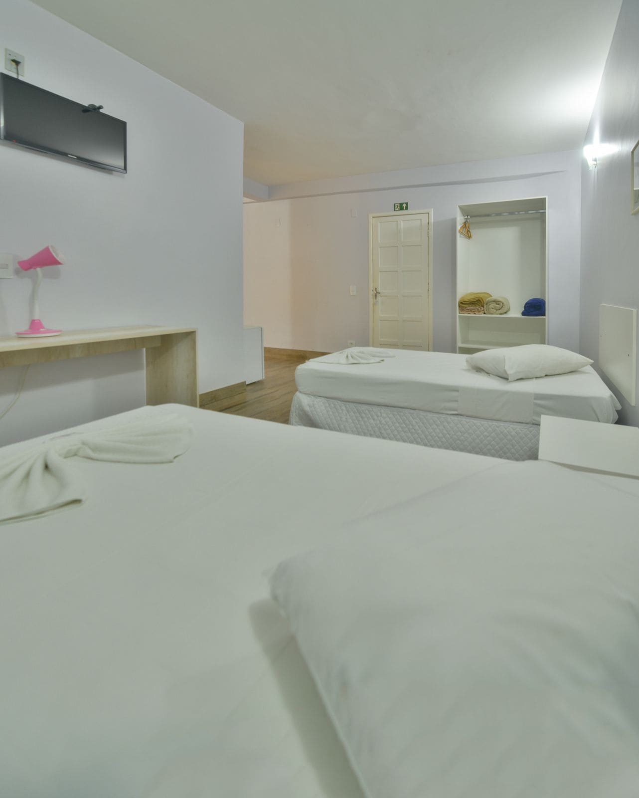Quarto de hotel com duas camas, uma escrivaninha com luminária, um guarda-roupa e uma televisão na parede.