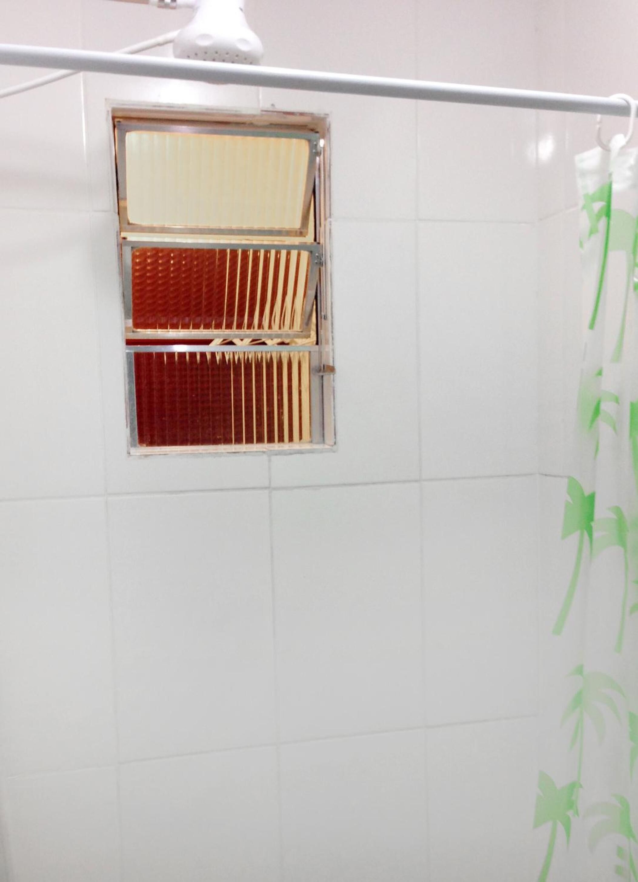 Casa de banho com cortina de chuveiro com padrões de palmeiras verdes, uma janela com moldura metálica e paredes revestidas de azulejos brancos.