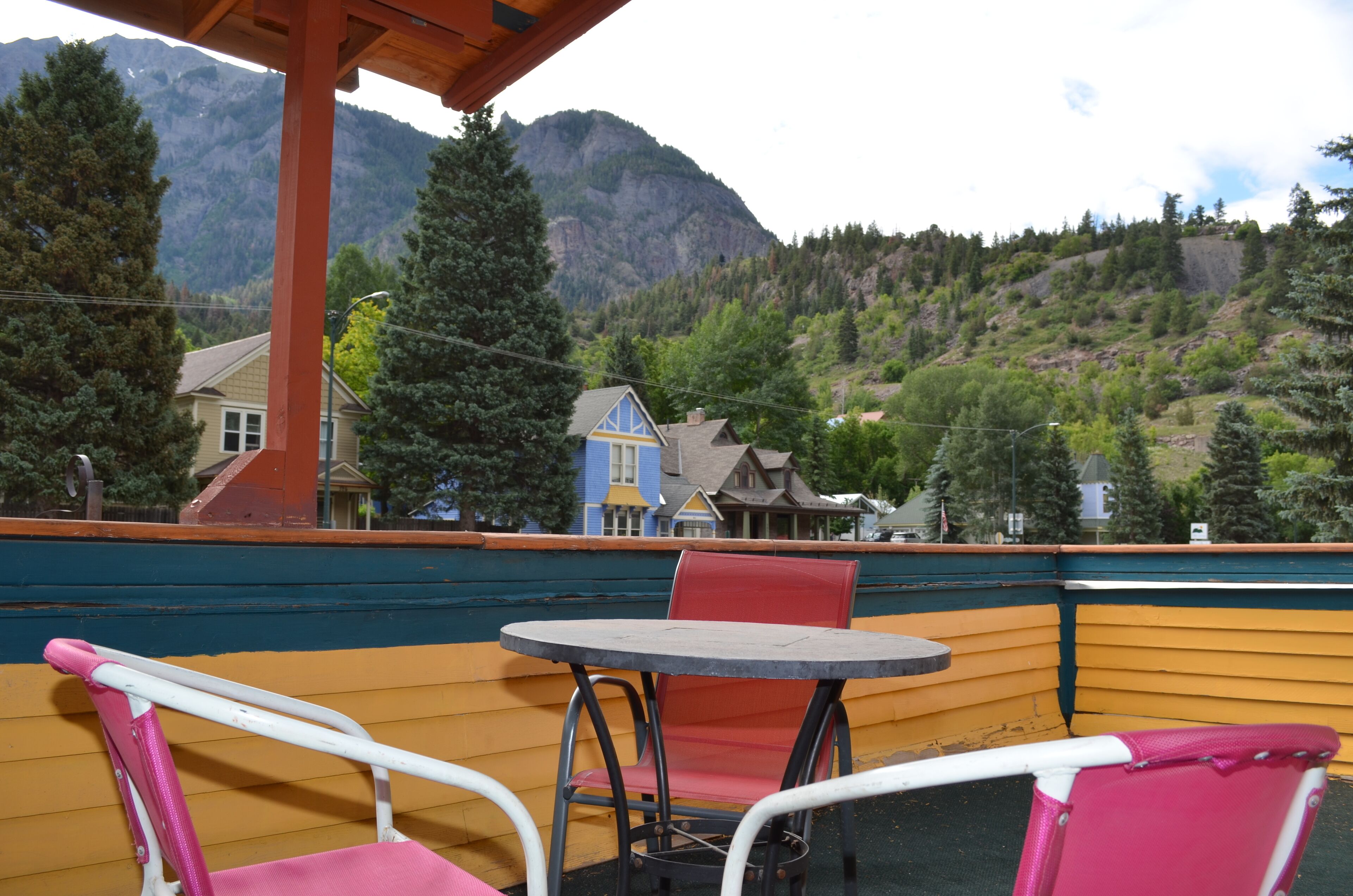 Um terraço com cadeiras coloridas e uma mesa, com vista para uma paisagem montanhosa com casas.
