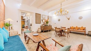 Uma sala de estar ampla com uma mesa de jantar de madeira, um sofá azul e um banco de madeira. Há um candelabro, uma cabeça de veado fixada na parede e um armário de madeira.