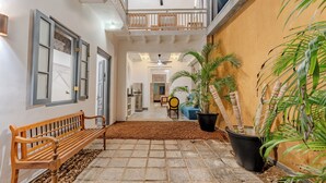 Um corredor com um banco de madeira, plantas em vasos e um chão de azulejos que leva a um quarto com ventoinha de teto.