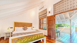 Quarto com cama de madeira, um banco verde, mesa de cabeceira e janela com cortinas estampadas.