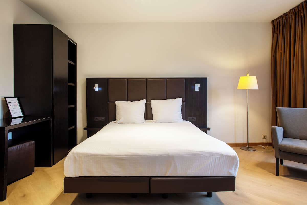 Quarto de hotel bem organizado, com uma cama grande, uma escrivaninha com cadeira, um abajur e um guarda-roupa.
