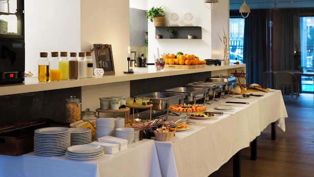 Buffet com diversos alimentos e bebidas, uma máquina de café e área de refeições com cadeiras e mesa.