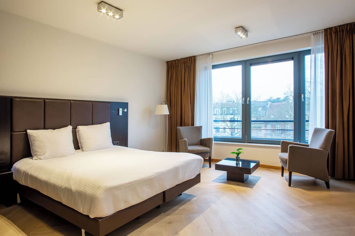 Quarto de hotel com uma cama grande, duas poltronas, uma mesinha e uma janela com cortinas.