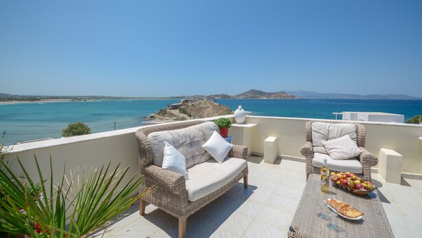 Um terraço com móveis de vime, uma mesa com comida e bebida, e vista para o mar e as montanhas.