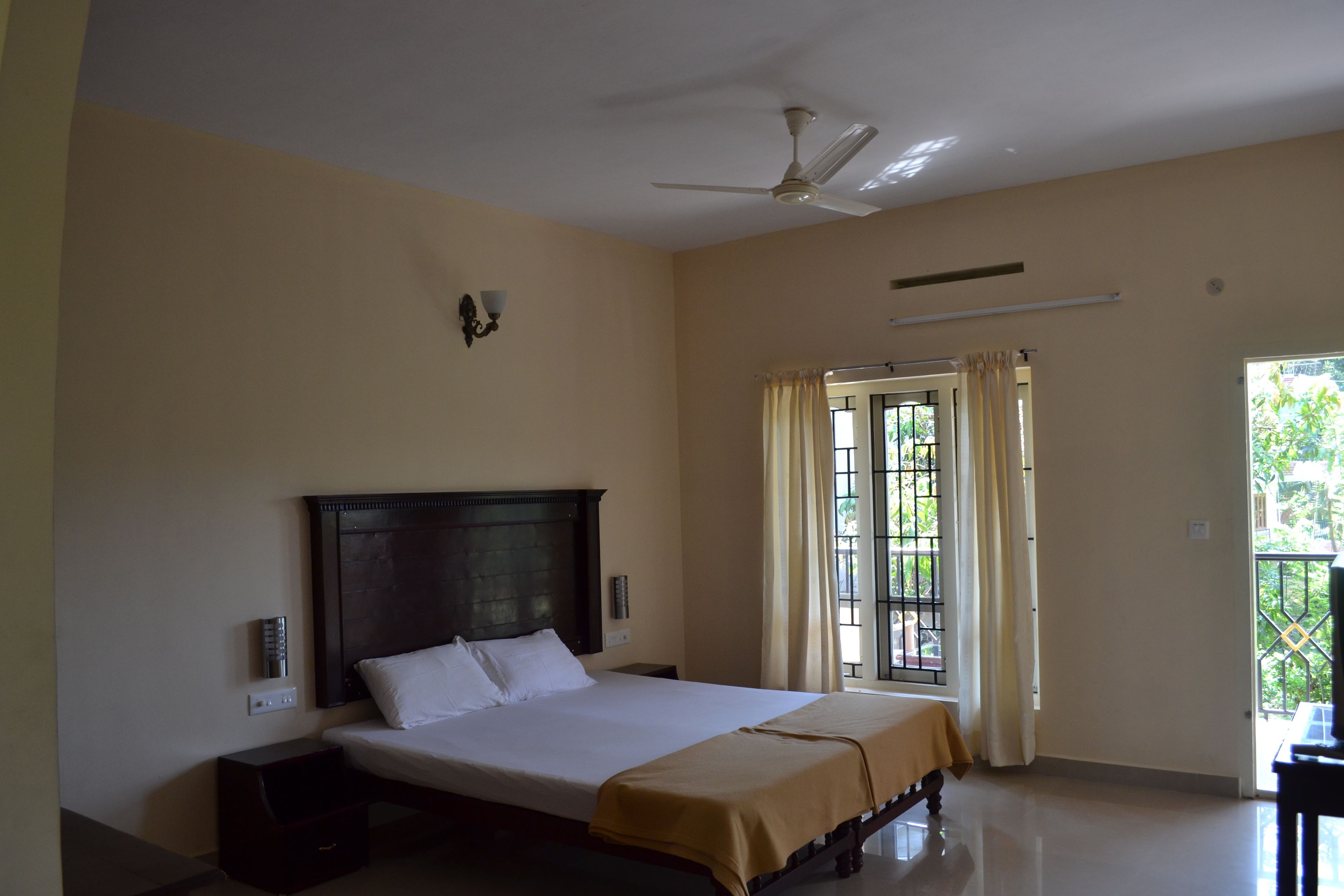 Quarto com cama, ventilador de teto, janela com cortinas e vista para o verde lá fora.