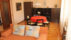 Uma sala de estar acolhedora com um sofá, duas almofadas iguais, uma mesa de jantar vermelha com quatro cadeiras e uma área de cozinha ao fundo.