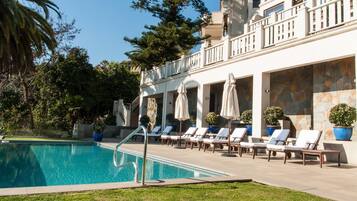 Piscina com espreguiçadeiras e guarda-sóis, cercada por vegetação e um edifício com paredes de pedra e varandas.