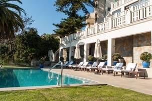 Piscina com espreguiçadeiras e guarda-sóis, cercada por vegetação e um edifício com paredes de pedra e varandas.