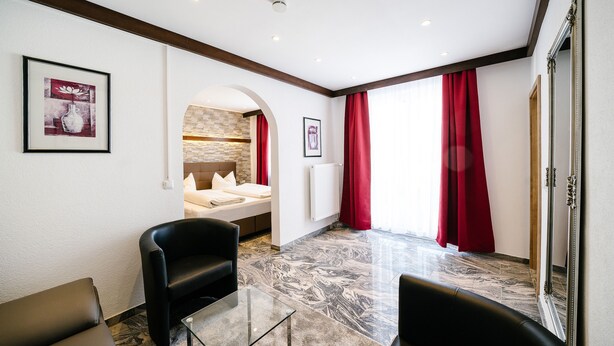 Quarto de hotel com uma cama, área de estar com cadeiras, mesa de vidro, lareira e cortinas vermelhas.