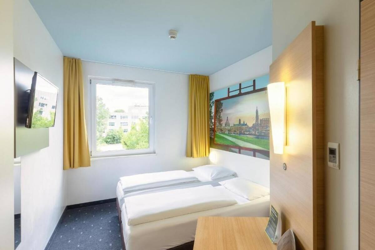 Quarto de hotel com cama, escrivaninha, janela com cortinas, quadro na parede e televisão fixada na parede.
