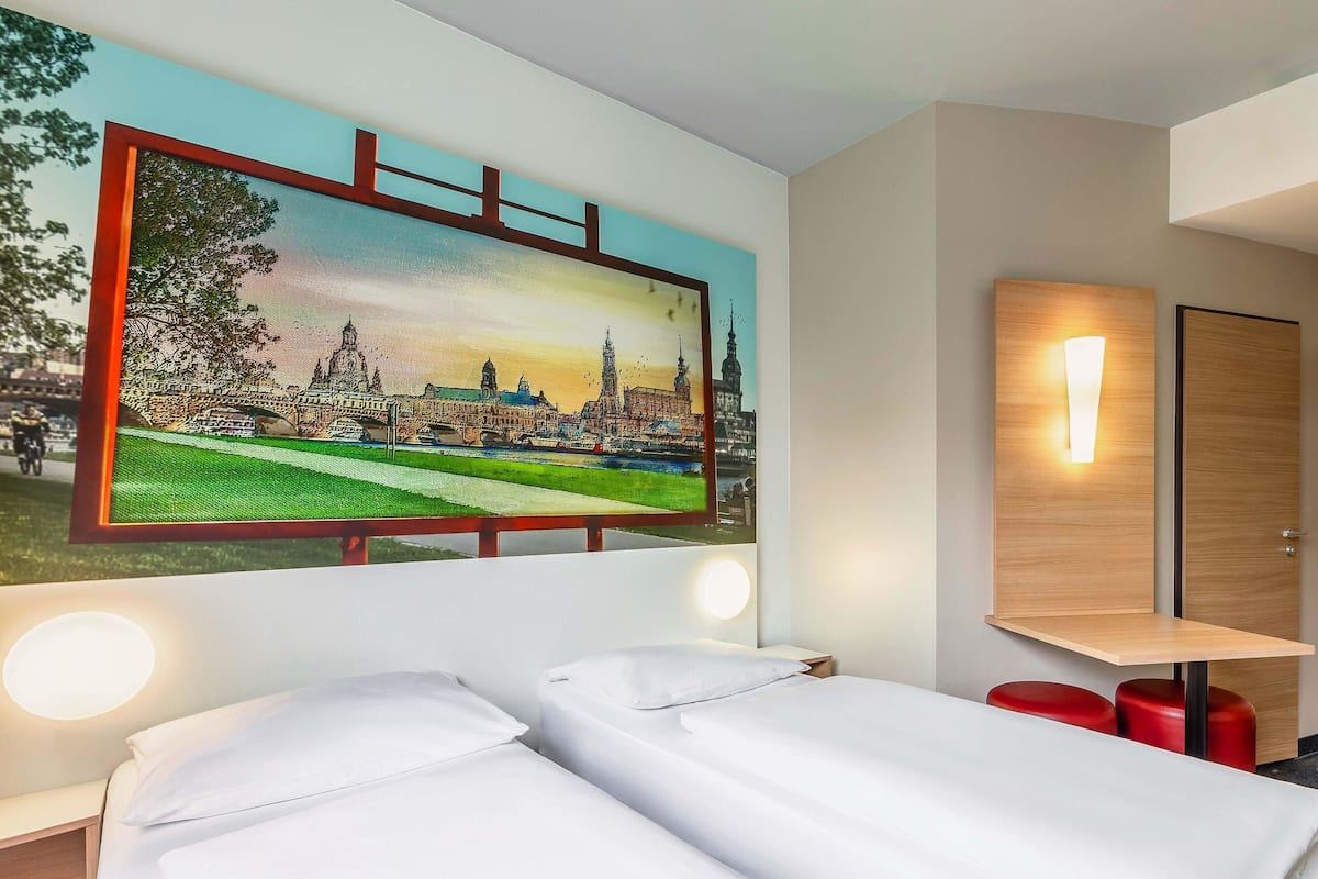Quarto de hotel com uma cama, uma mesa de cabeceira, uma luminária na parede e uma pintura de uma paisagem urbana.