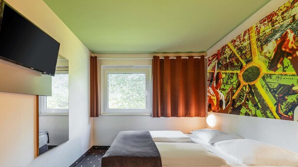 Quarto de hotel com cama, escrivaninha, televisão, janela com cortinas e uma grande pintura abstrata na parede.