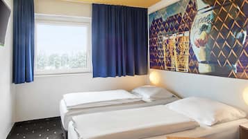 Quarto de hotel com duas camas, janela com cortinas, luminária na parede e vista para a cidade.