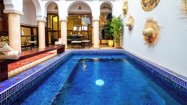 Área de piscina com borda em azulejo, espaço de estar com cadeiras e mesa, e detalhes decorativos na parede.