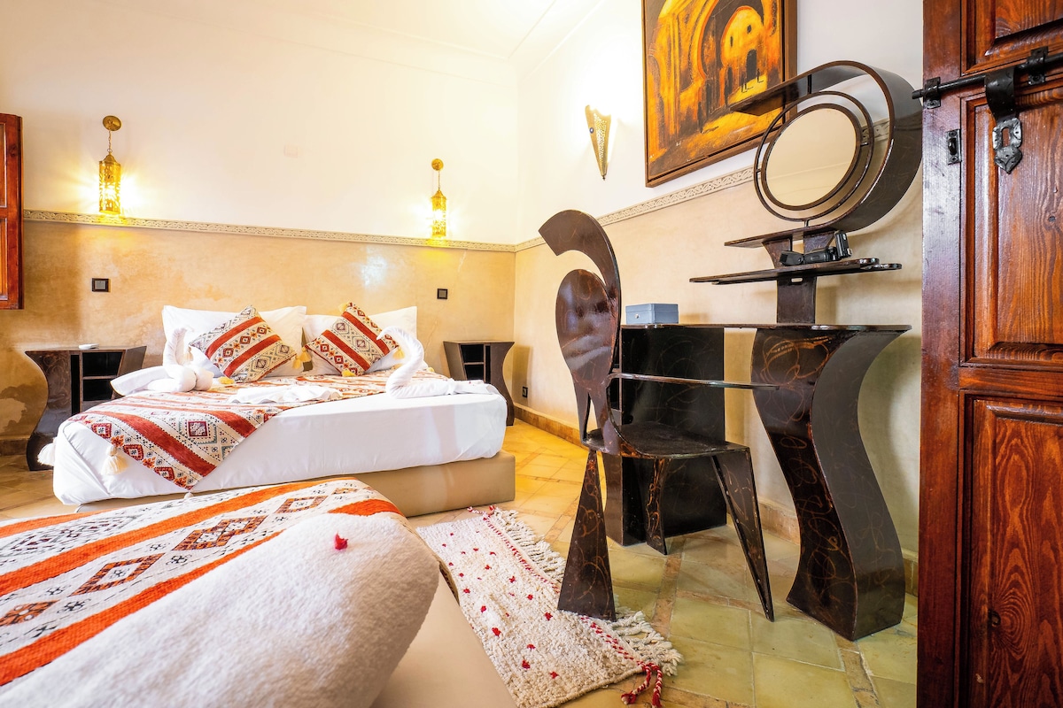 Quarto de hotel com cama, mesas de cabeceira, escrivaninha, cadeira, espelho e um quadro na parede.