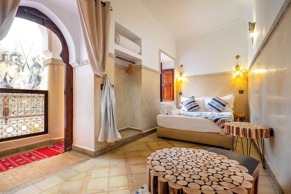 Quarto de hotel com cama, mesa redonda pequena, janela com cortinas e vista para o exterior.