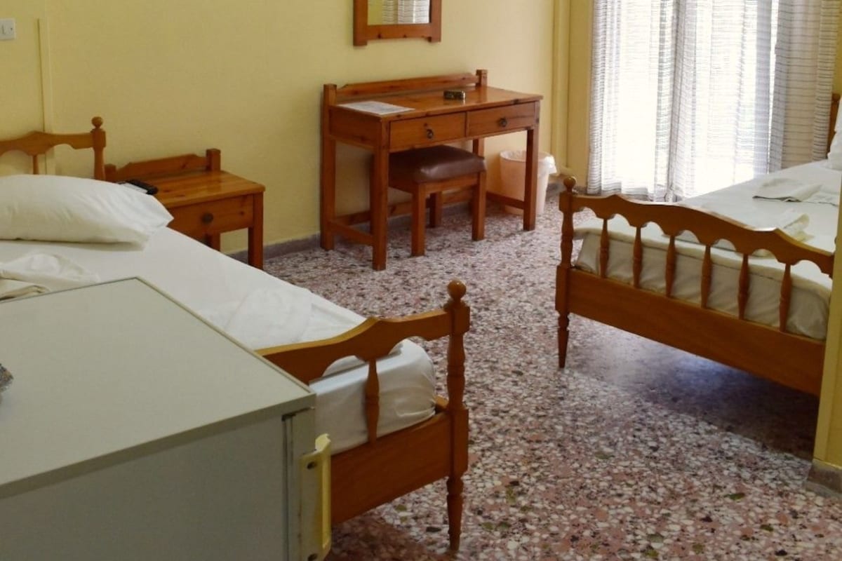Quarto com duas camas de madeira, uma escrivaninha de madeira, uma cadeira, um espelho e um ar condicionado.