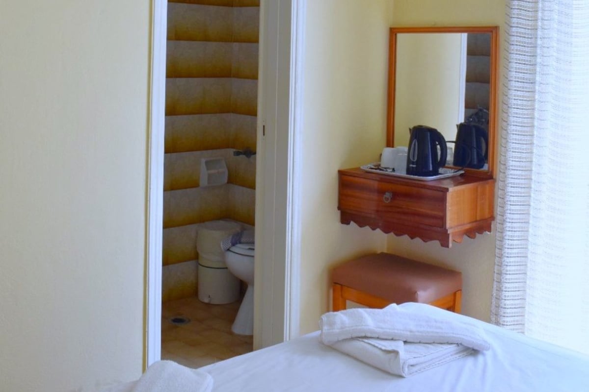 Quarto de hotel com cama, televisão na parede, banheiro com cortina de chuveiro e pequena mesa de madeira com chaleira e xícaras.