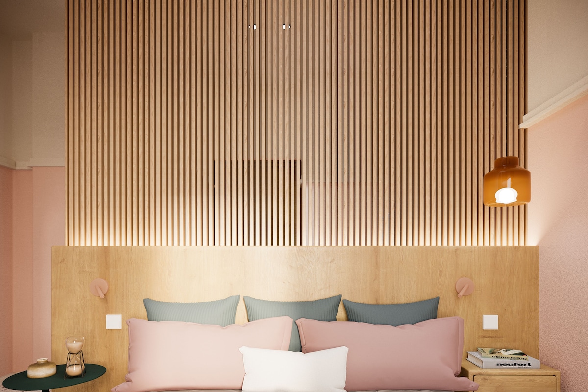 Quarto com cabeceira de ripas de madeira, cama com roupa de cama rosa, duas mesas de cabeceira com abajures e uma mesinha de cabeceira com um livro.