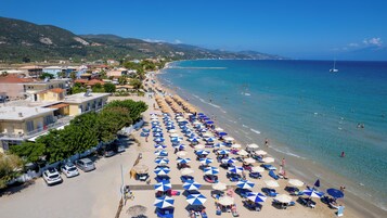 Uma vila costeira com uma praia repleta de guarda-sóis e espreguiçadeiras, mar azul translúcido e edifícios ao longo da orla.