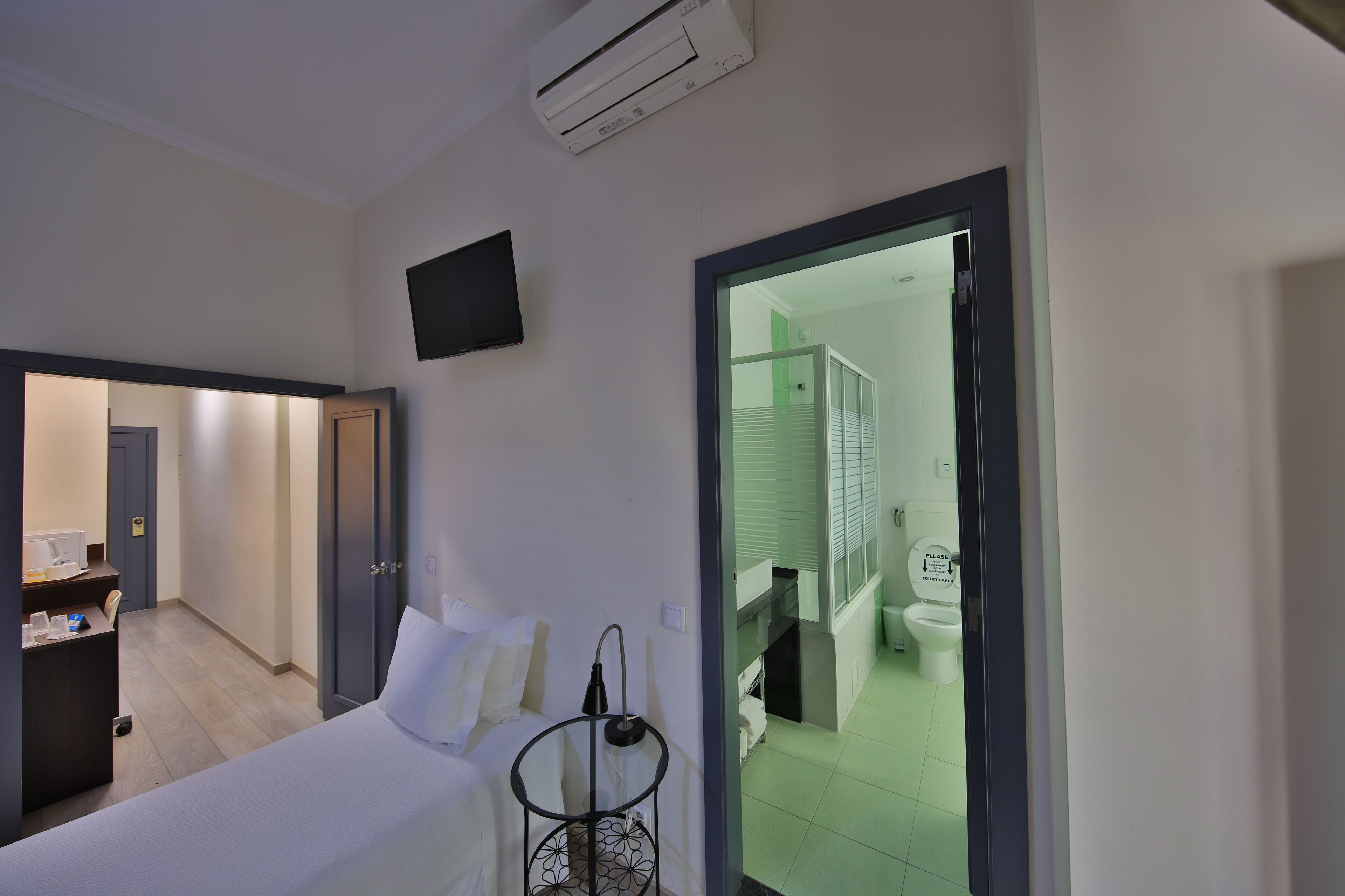 Quarto de hotel com cama, mesa de cabeceira, luminária e vista para o banheiro através de uma porta de vidro.