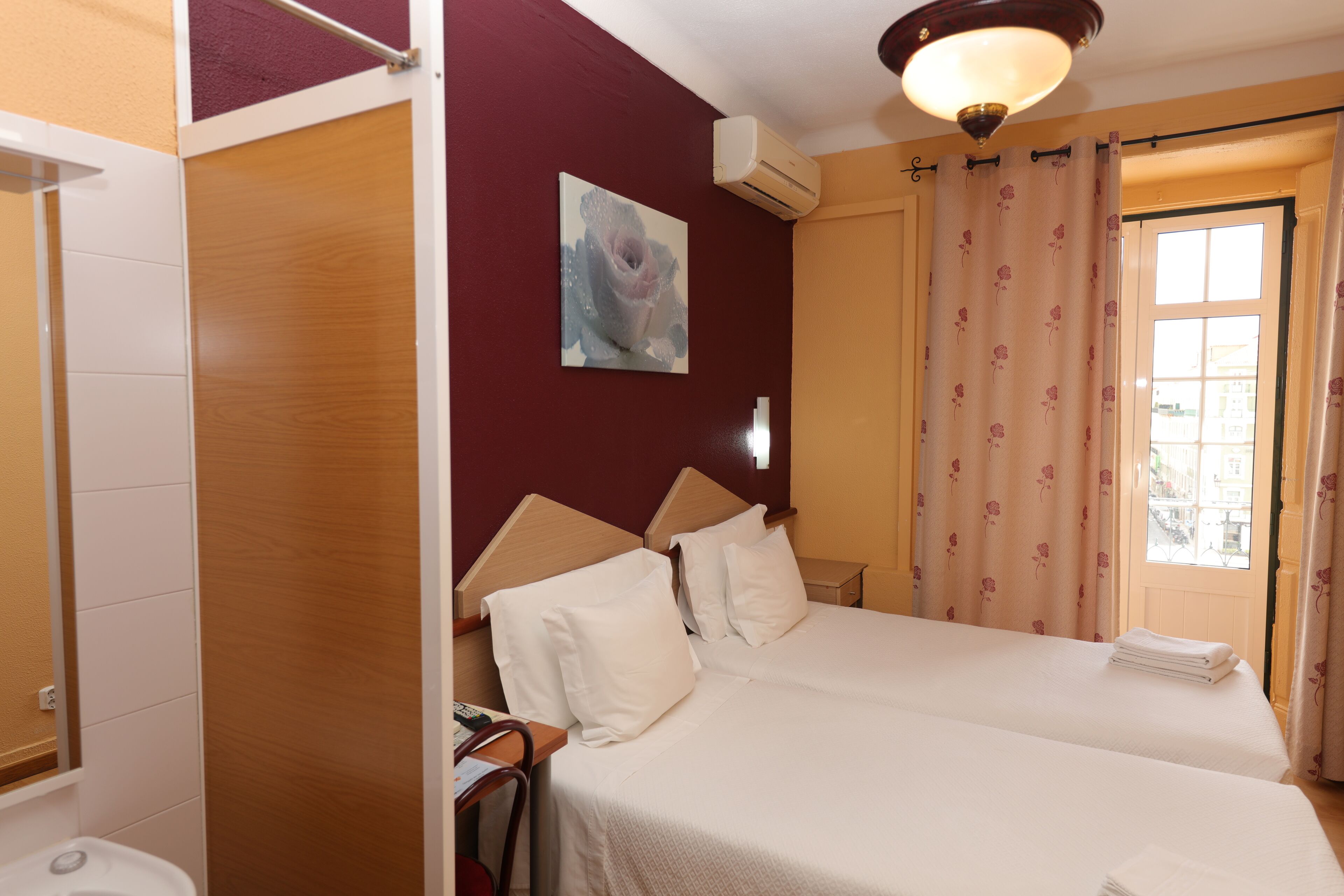 Quarto de hotel com duas camas, banheiro e janela com cortinas florais.