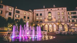 Um edifício histórico com uma placa indicando 'PIAZZA HOTEL' e uma fonte com vários jatos de água.