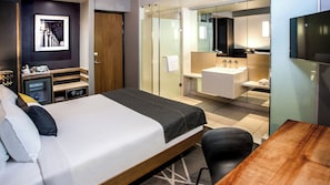 Quarto de hotel com cama, escrivaninha, espelho, banheiro com pia e chuveiro, e um pequeno armário.