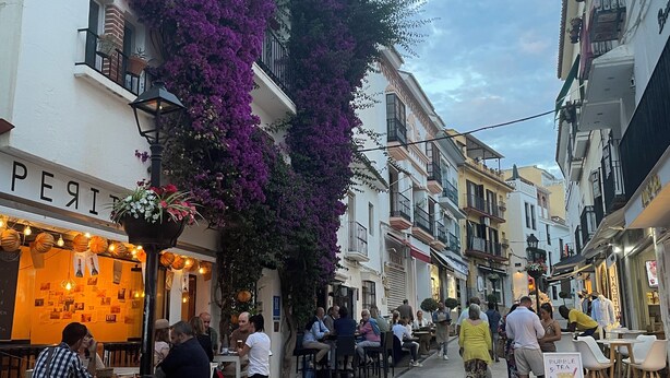 Rua estreita com cafés e lojas, com mesas ao ar livre e trepadeiras roxas florescendo.