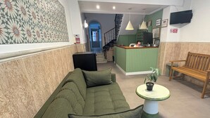 Um sofá verde com uma mesinha e uma planta, um banco de madeira, um balcão de recepção e uma escadaria que leva a um andar superior.