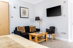 Quarto de hotel moderno com cama, sofá, mesa de centro, poltrona, televisão e luminária.