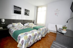 Quarto de hotel com duas camas, uma escrivaninha, uma cadeira, um radiador e uma janela com cortinas.