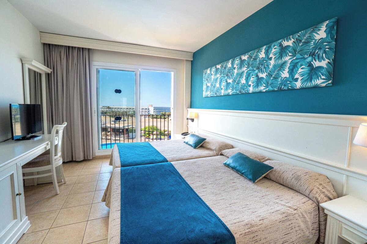 Quarto de hotel com duas camas, uma mesa, televisão e varanda com vista.