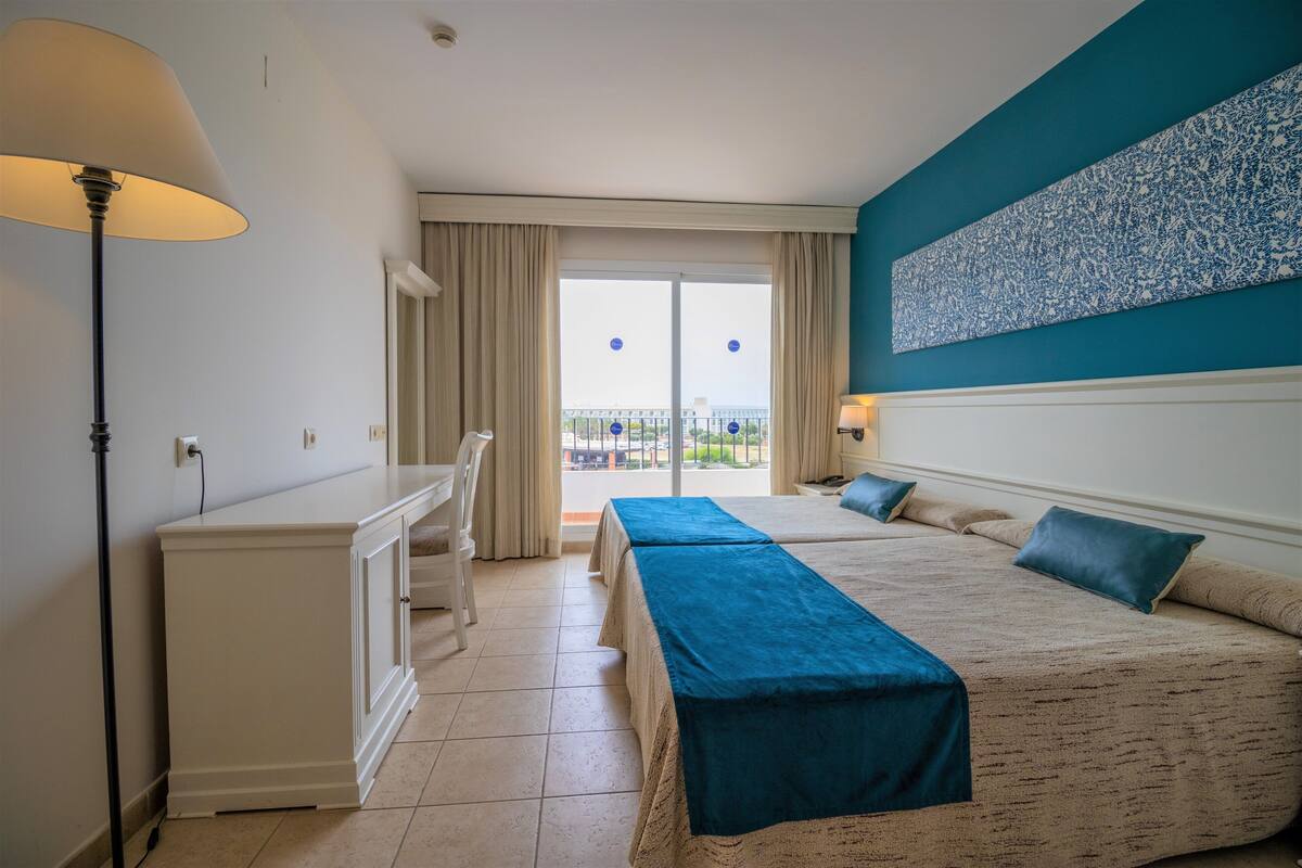 Quarto de hotel com cama, escrivaninha e cadeira. Há vista para o exterior por uma janela grande.