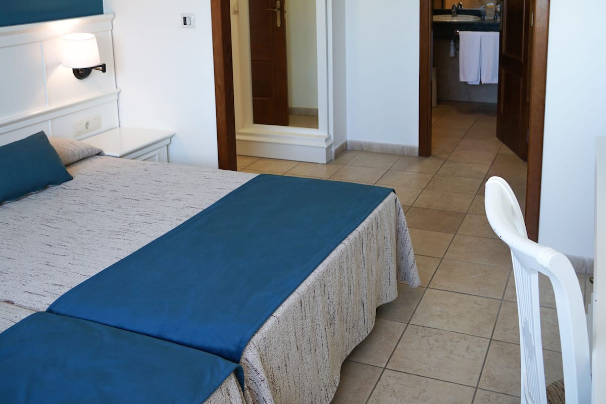Quarto com cama, mesa de cabeceira e uma cadeira. Um banheiro é visível através de uma porta aberta.