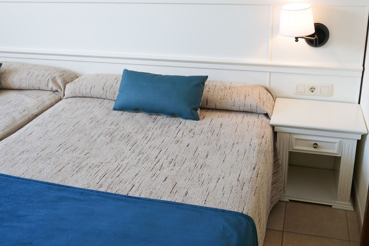 Quarto de hotel com uma cama grande, mesa de cabeceira, luminária fixada na parede e uma pintura abstrata azul na parede.
