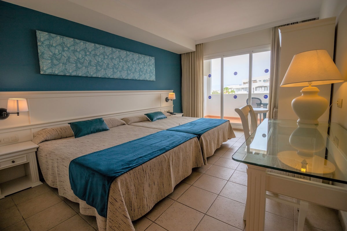Quarto de hotel com duas camas, uma mesa de vidro, uma cadeira e vista para a varanda externa.