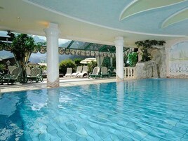 Piscina interior com água azul cristalina, rodeada por espreguiçadeiras e plantas em vasos.