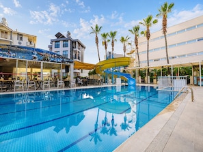 Piscina com escorregador, palmeiras e um edifício com restaurante.