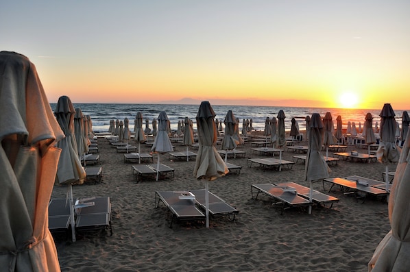 Uma praia com fileiras de espreguiçadeiras e guarda-sóis, alguns cobertos com panos, e um pôr do sol ao fundo.