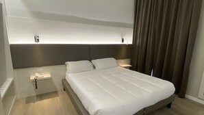 Quarto de hotel moderno com uma cama grande, mesas de cabeceira e um criado-mudo embutido.