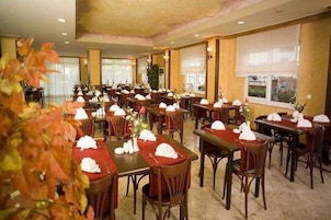 Uma sala de jantar com várias mesas dispostas com toalhas brancas e jogos americanos vermelhos, com um arranjo floral central.
