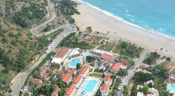 Vista aérea de um resort à beira-mar, com várias piscinas, uma praia e uma estrada sinuosa.