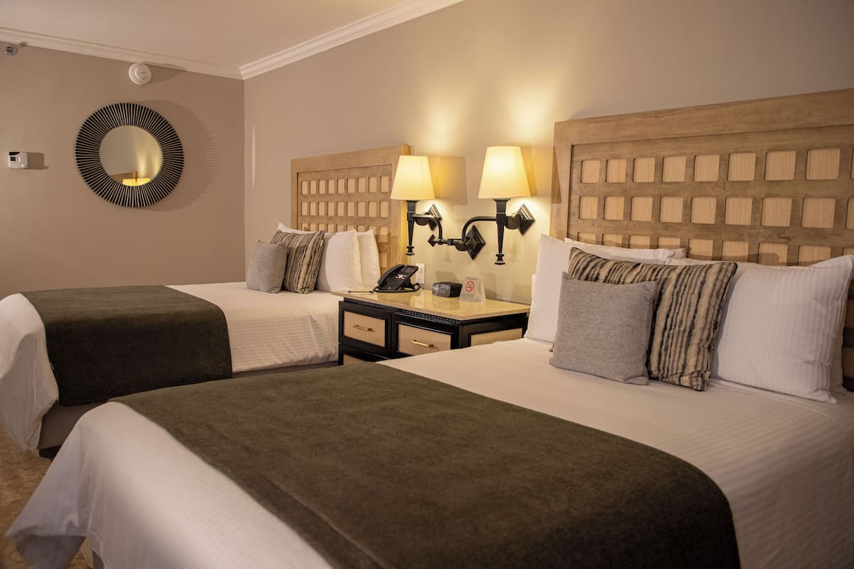 Quarto de hotel com duas camas, uma mesa de cabeceira com abajur, um espelho e uma cabeceira de madeira.